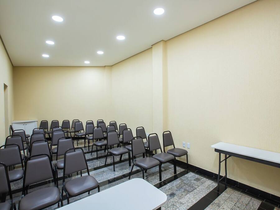 Uma sala de conferências com fileiras de cadeiras, uma mesa branca e uma tela de projeção.