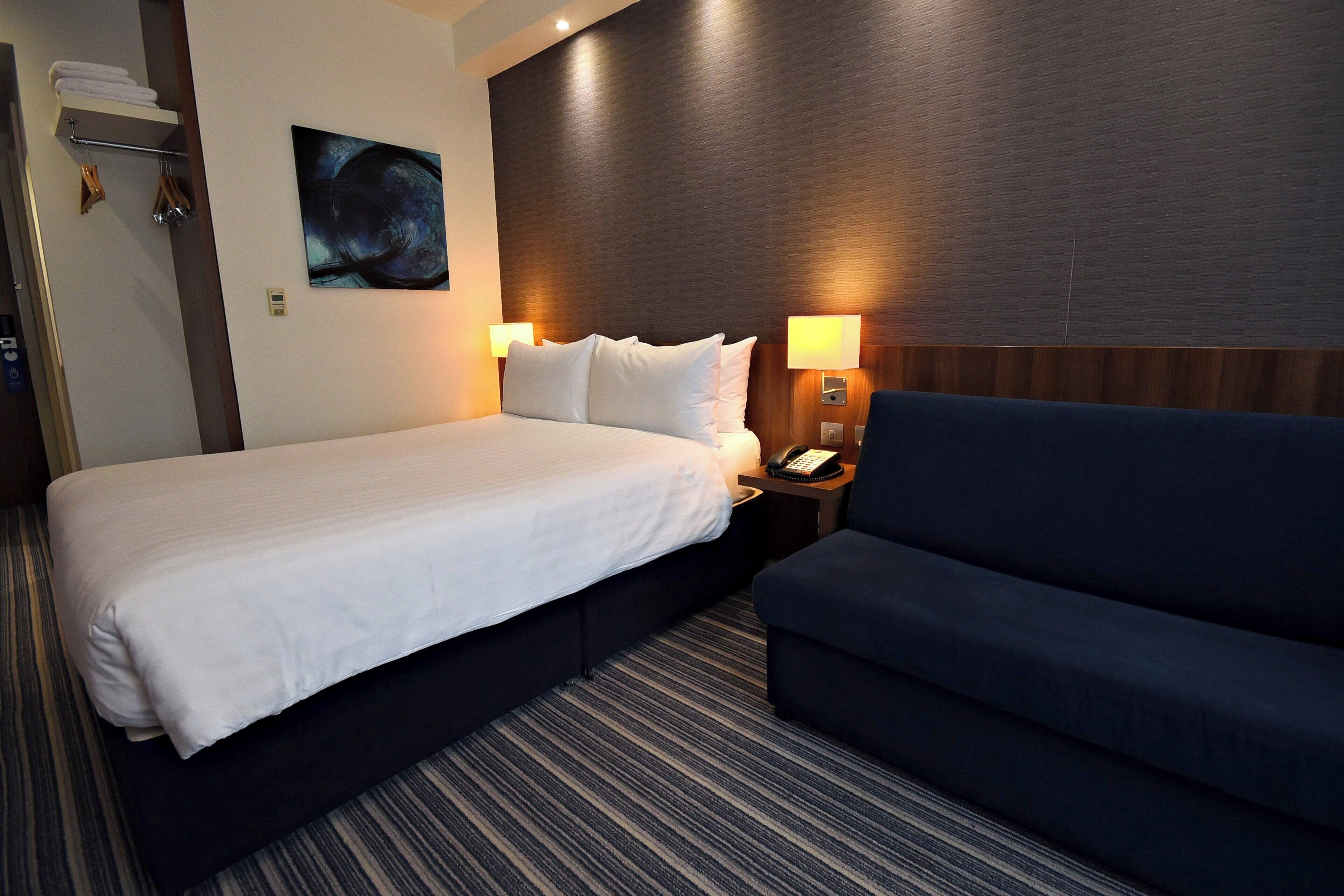 Quarto de hotel com cama, mesa de cabeceira com abajur, telefone e uma obra de arte na parede.