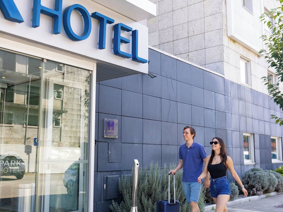 Entrada de um hotel com porta de vidro, placa com a palavra 'HOTEL' e uma pessoa com mala.