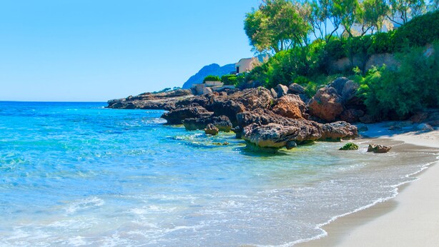 Uma praia com águas azul-claras, afloramentos rochosos e vegetação.