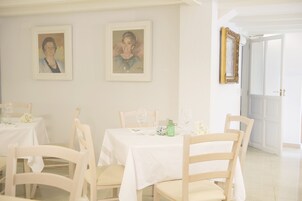 Uma área de refeições com mesas e cadeiras brancas, retratos nas paredes e uma porta que dá para outro cômodo.
