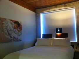 Quarto com cama, mesa de cabeceira, luminária na parede e janela com persianas.