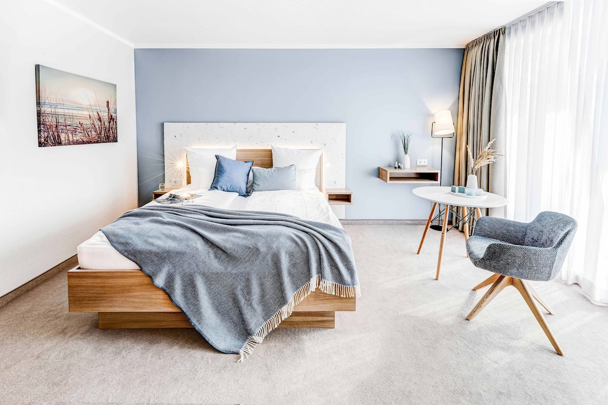Quarto de hotel moderno com cama, mesas de cabeceira, uma cadeira e uma pequena mesa com abajur.