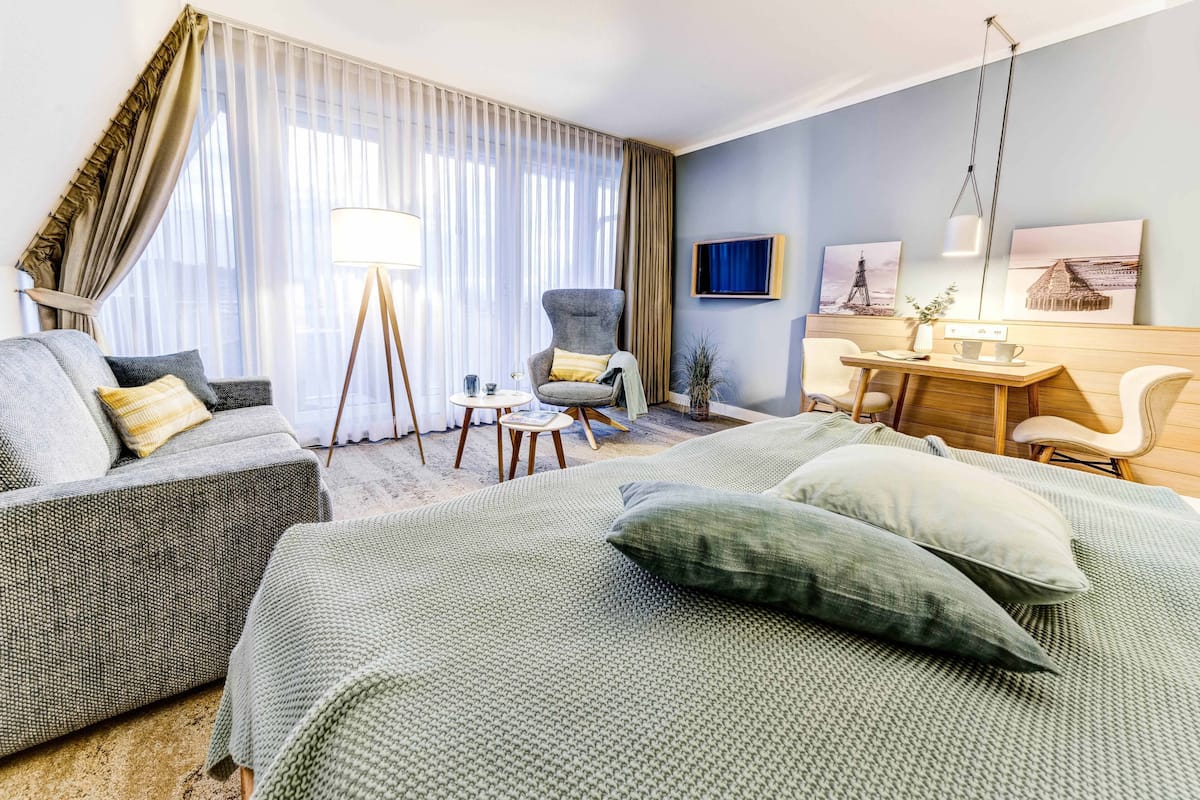 Quarto de hotel com cama, sofá, escrivaninha e cadeira. Há uma luminária, uma mesinha e uma planta.