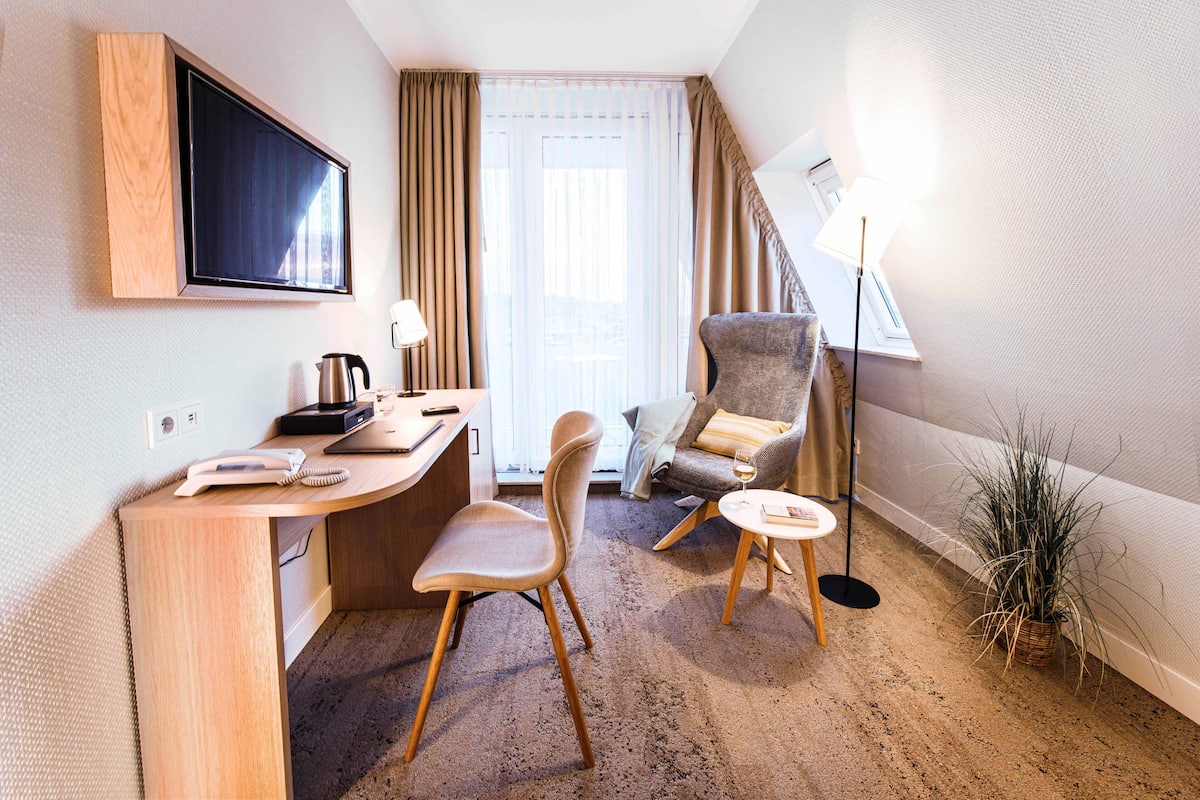 Quarto de hotel com escrivaninha, duas cadeiras, televisão, luminária e janela com cortinas.