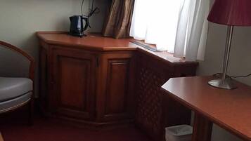 Quarto com televisão de tela plana na parede, um armário de madeira, uma cadeira, uma mesa pequena com abajur e uma janela com cortinas.