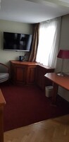 Quarto com televisão de tela plana na parede, um armário de madeira, uma cadeira, uma mesa pequena com abajur e uma janela com cortinas.
