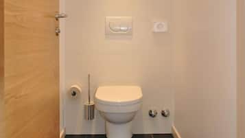 Um banheiro pequeno e moderno, com vaso sanitário branco, suporte para papel higiênico e botão de descarga na parede.