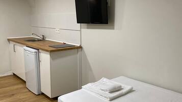 Quarto pequeno e limpo, com uma cama, escrivaninha com pia e televisão fixada na parede.