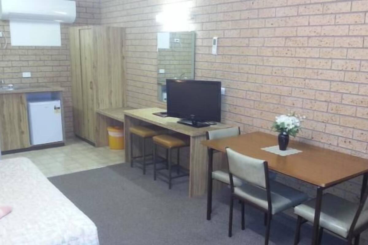 Quarto de hotel compacto com cozinha compacta, uma cama, uma mesa com computador e uma pequena mesa de jantar.