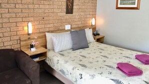 Quarto com parede de tijolos, uma cama com colcha estampada, duas almofadas, uma mesa de cabeceira com abajur, cabeceira de madeira e um quadro na parede.