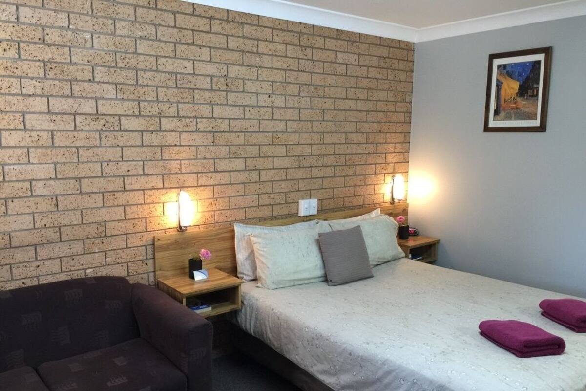 Quarto de hotel com parede de tijolos, cama com almofadas, mesa de cabeceira com abajur, uma cadeira e um quadro na parede.