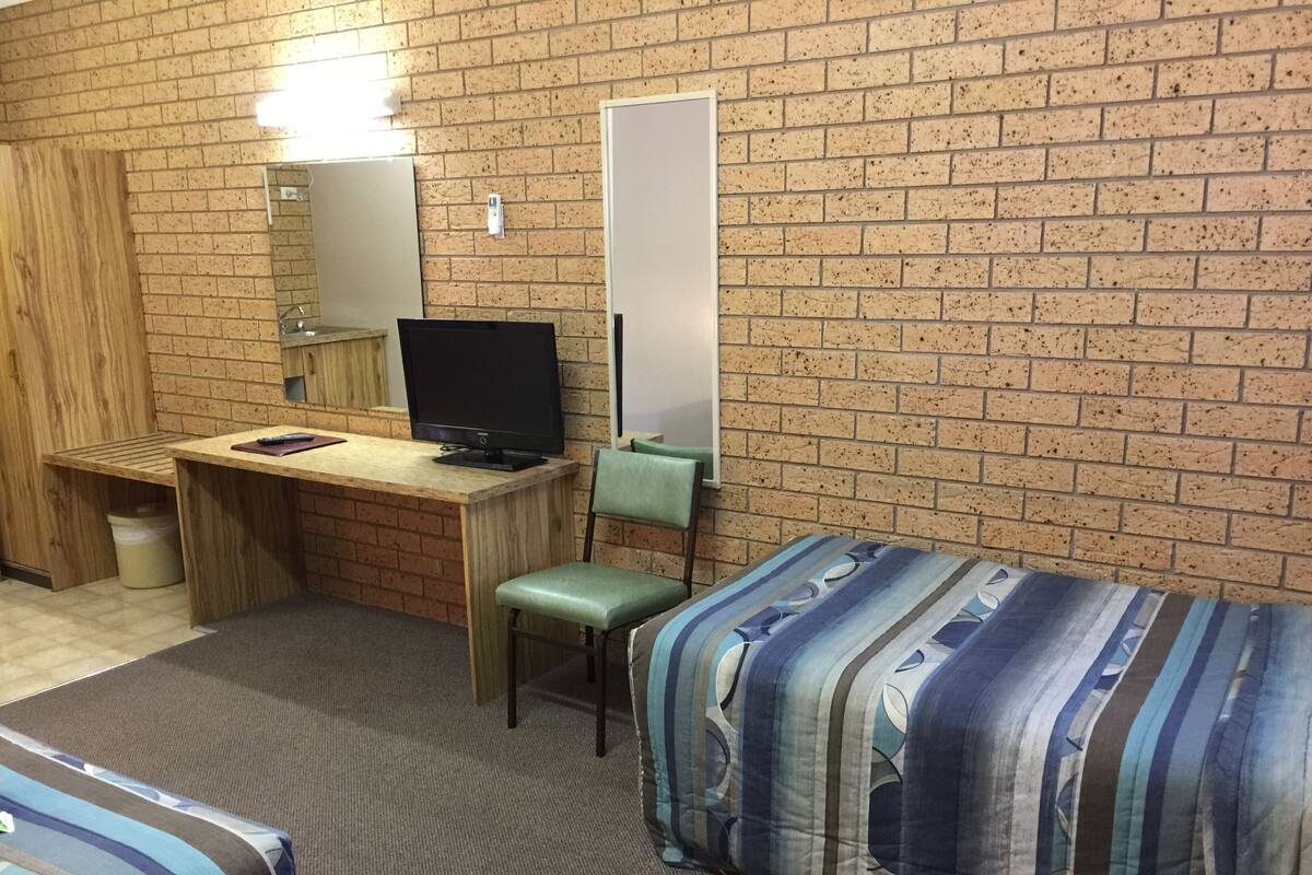 Quarto com parede de tijolos, uma escrivaninha com computador, uma cadeira e duas camas com roupa de cama listrada.
