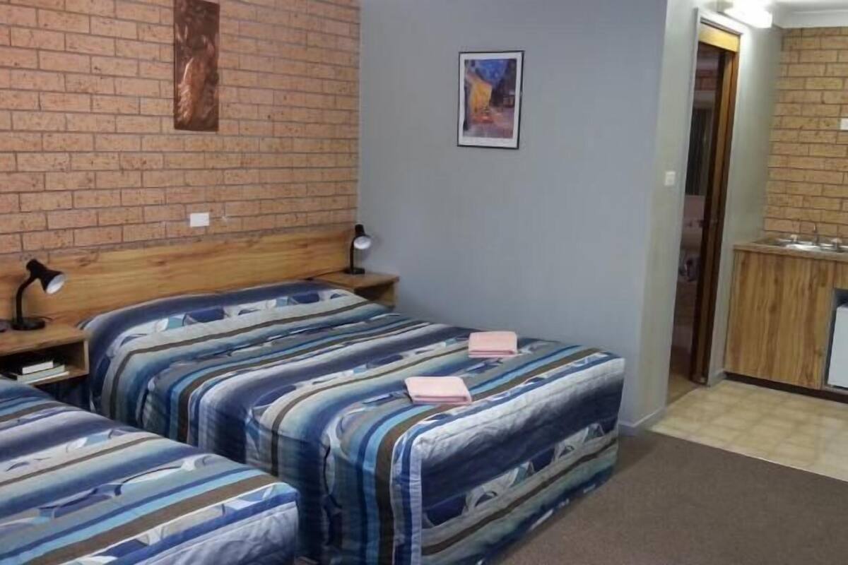 Quarto com duas camas, cabeceira de madeira, uma pequena escrivaninha com luminária, um quadro na parede e um espaço com cozinha compacta.