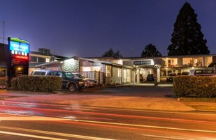 Hotel com área de recepção, estacionamento e uma grande árvore ao fundo.
