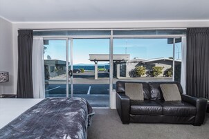 Quarto de hotel com cama, sofá de couro e uma grande porta de vidro deslizante que dá para um estacionamento e um edifício com vista para o mar.