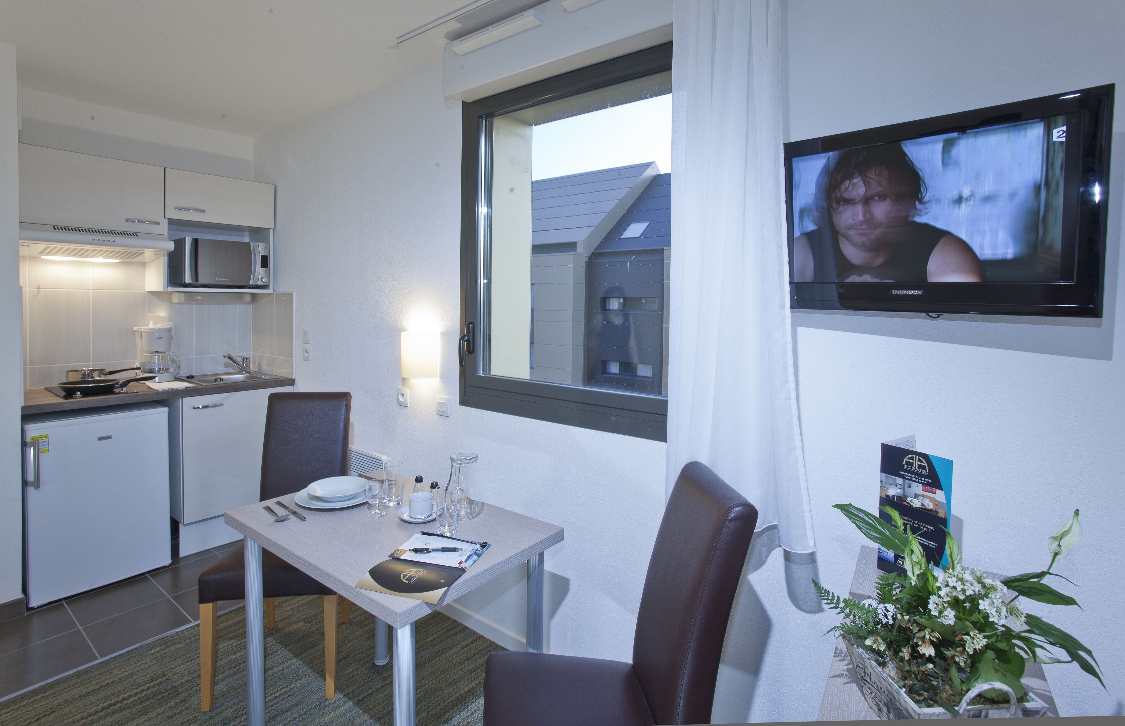 Quarto de hotel moderno com cozinha compacta, área de refeições, TV na parede e janela com vista para o exterior.
