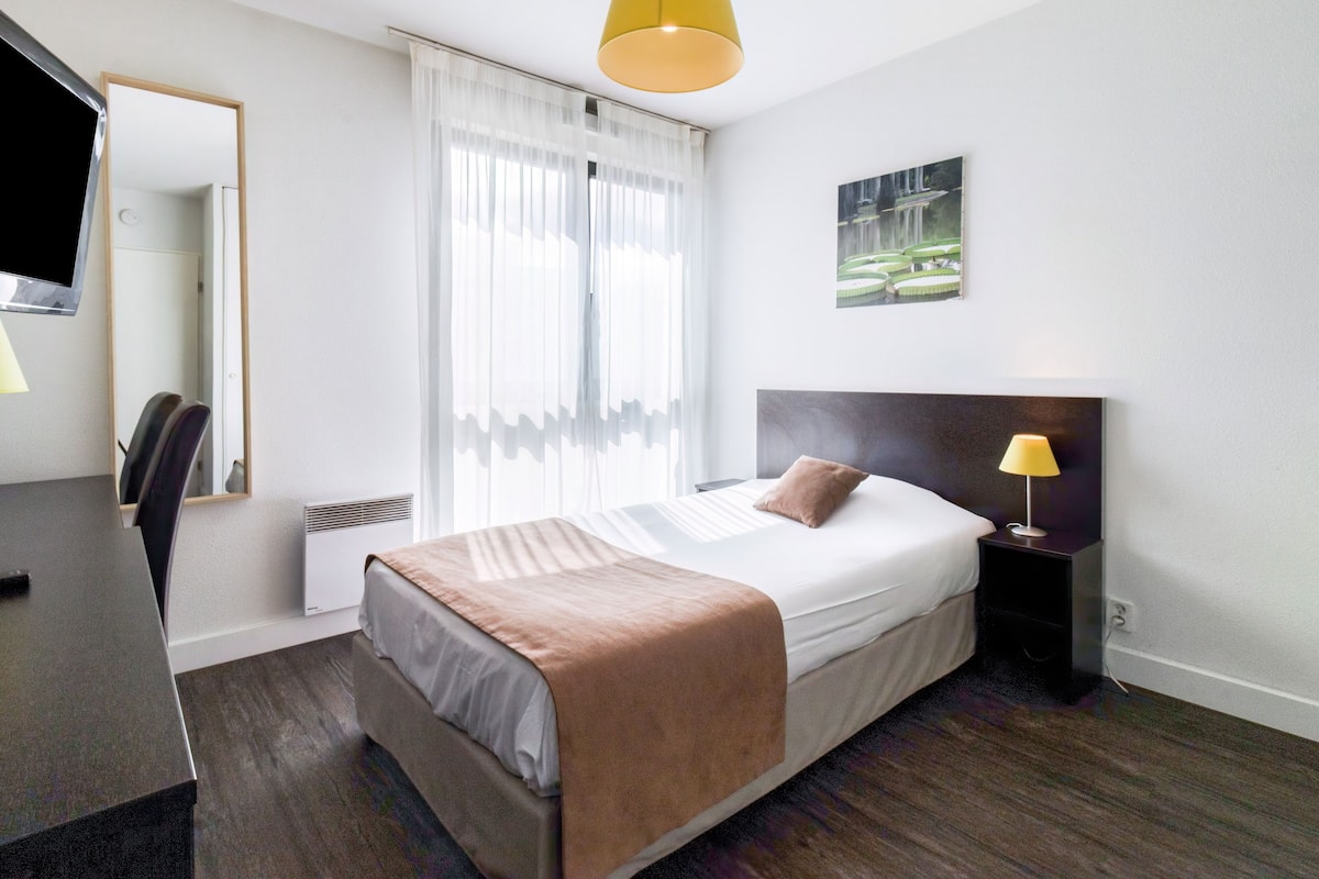 Quarto de hotel com cama, escrivaninha, cadeira, mesa de cabeceira com abajur, janela com cortinas e um quadro na parede.