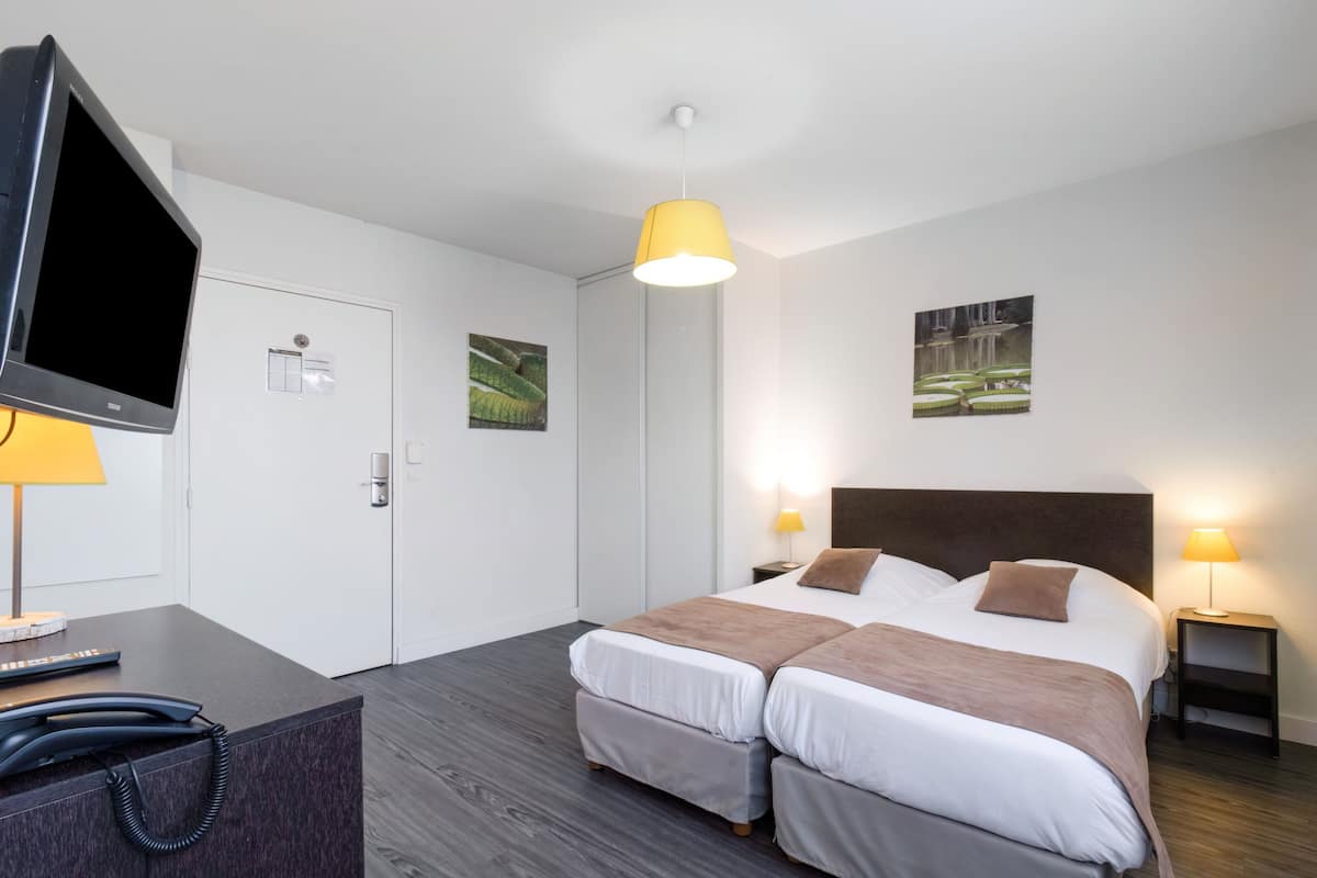 Quarto de hotel com cama, luminárias de cabeceira, escrivaninha com telefone, televisão e obras de arte nas paredes.