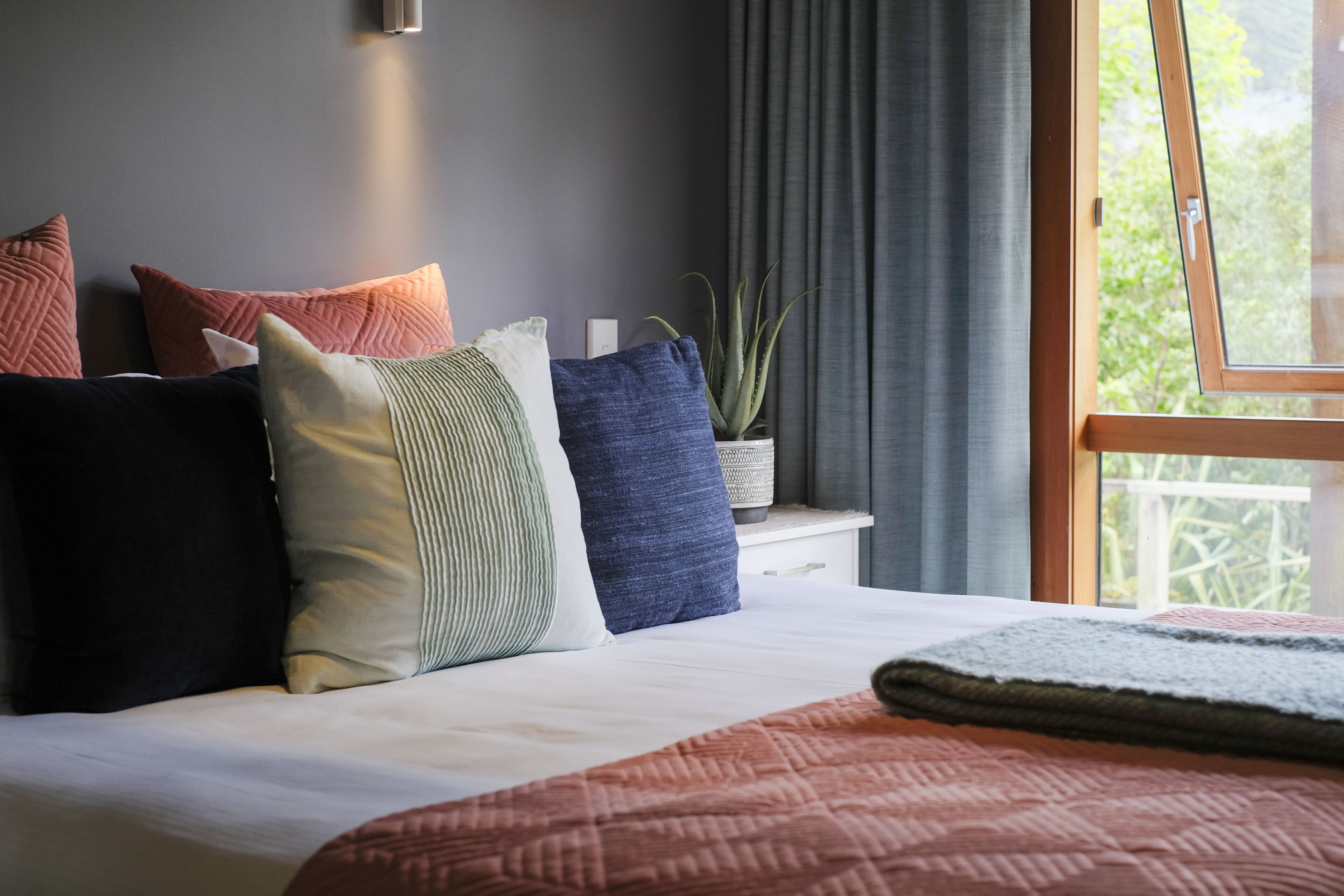 Cama bem arrumada com várias almofadas, um vaso de planta e uma janela com cortinas.