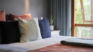Cama bem arrumada com várias almofadas, um vaso de planta e uma janela com cortinas.