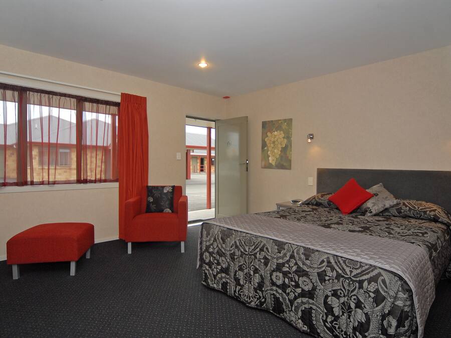 Quarto de hotel com cama, uma cadeira vermelha, um banquinho pequeno, janela com cortinas translúcidas e porta que dá para uma área externa.