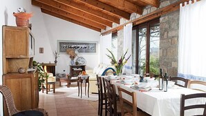 Uma área de refeições com mesa de madeira posta para quatro pessoas, rodeada por cadeiras. Uma parede de pedra e uma janela com cortinas são visíveis ao fundo.