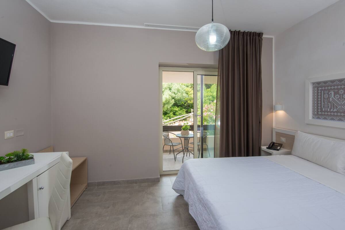 Quarto de hotel com uma cama, uma escrivaninha, uma cadeira, uma mesinha com uma planta, um telefone e vista para o exterior através de uma porta de vidro.