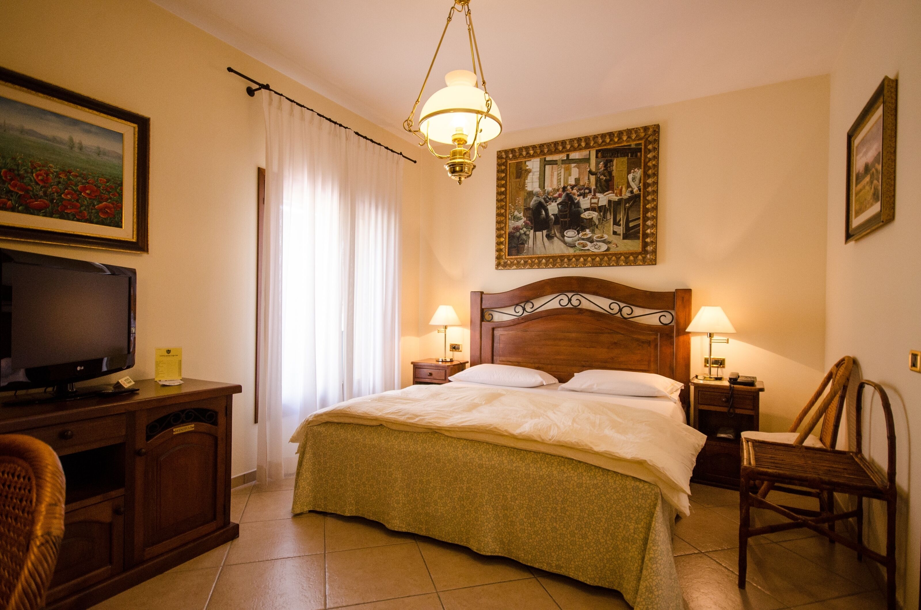 Quarto de hotel com uma cama, duas luminárias laterais, uma televisão sobre um armário de madeira, uma cadeira e um quadro na parede.