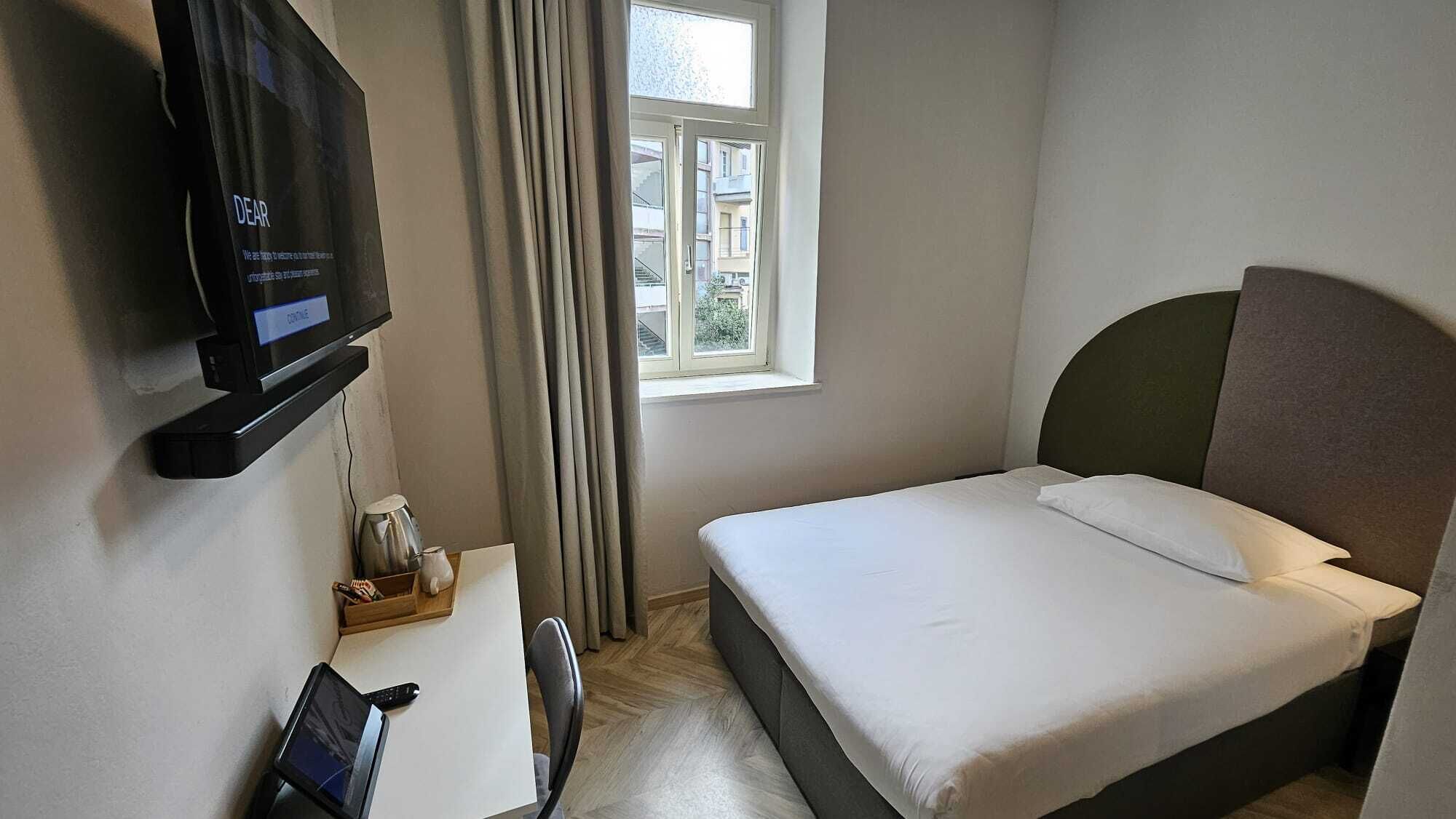 Quarto de hotel pequeno com uma cama, escrivaninha, televisão e janela, com vista para prédios.
