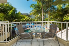 Varanda com mesa de vidro e duas cadeiras, com vista para uma área de piscina com guarda-sóis azuis e palmeiras.
