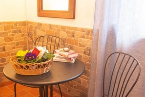 Um canto com parede de pedra, uma cesta de vime com flores, uma pilha de livros e uma cadeira de metal.