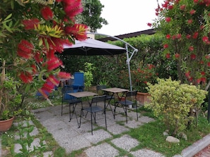 Um pátio externo com mesa e cadeiras, cercado por vegetação exuberante e plantas floridas.