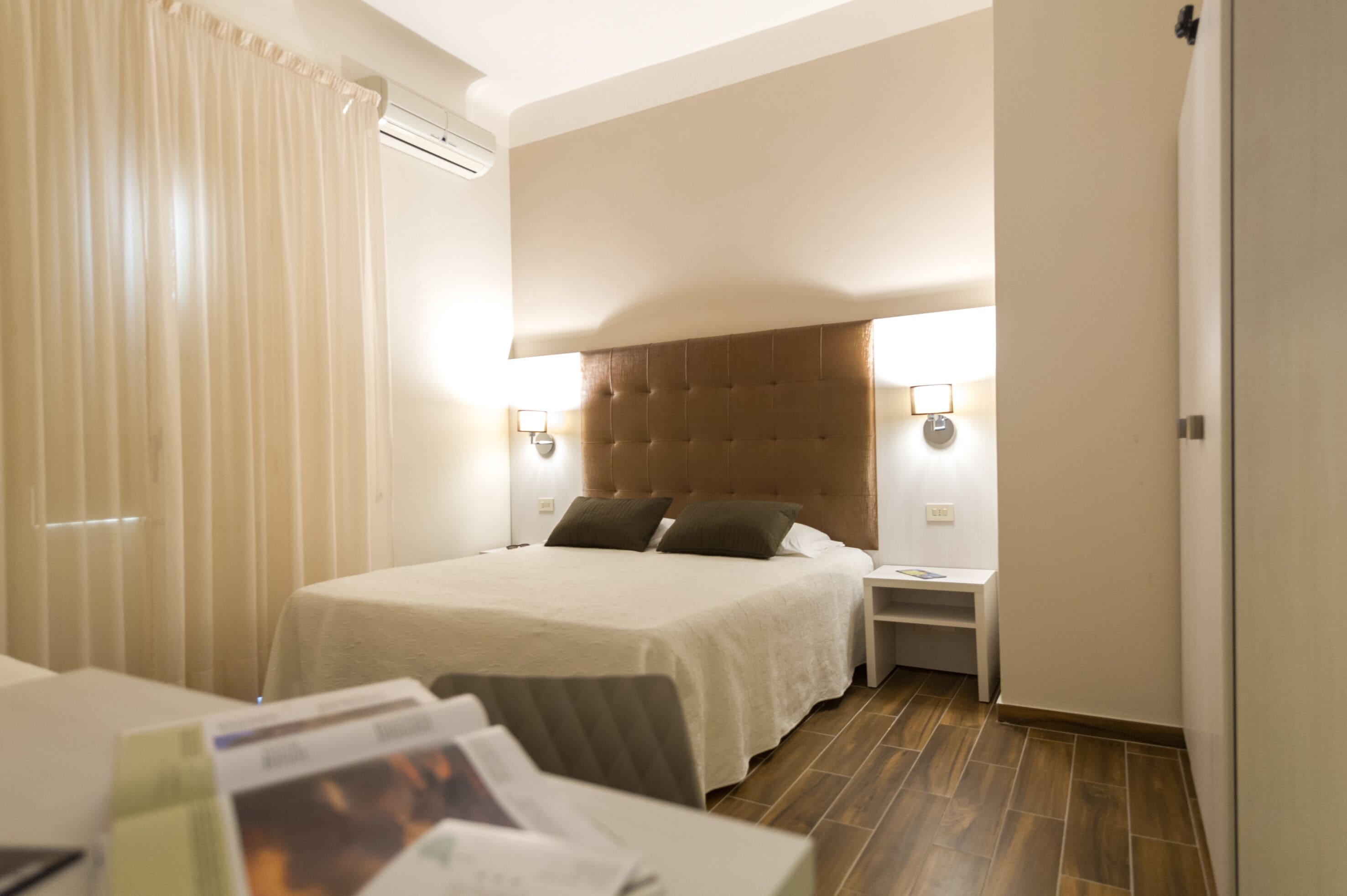 Quarto de hotel com uma cama grande, duas mesas de cabeceira e uma janela com cortinas.
