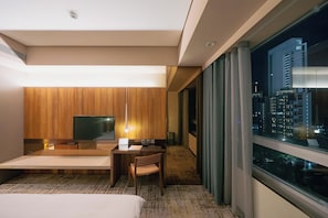 Quarto de hotel com cama, escrivaninha com cadeira, televisão e vista para a cidade à noite.
