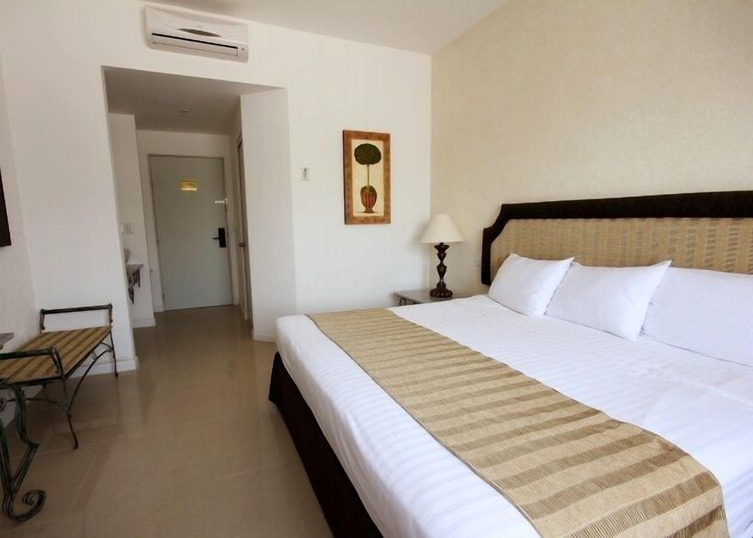 Quarto de hotel com uma cama grande, uma mesinha de cabeceira com abajur, uma cadeira e um quadro na parede.