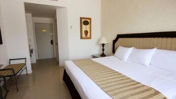Quarto de hotel com uma cama grande, uma mesinha de cabeceira com abajur, uma cadeira e um quadro na parede.