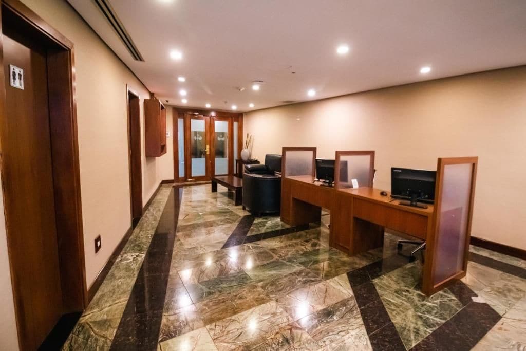 Corredor de hotel com piso de mármore, portas de madeira, balcão de recepção e área de estar.
