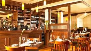 Um restaurante com mobiliário de madeira, um bar com prateleiras cheias de garrafas e um piano ao fundo.