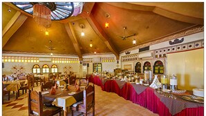 Uma sala de jantar ampla, com tetos altos, lustres e detalhes decorativos elaborados nas paredes. Mesas dispostas com toalhas vermelhas e diversos utensílios de jantar.