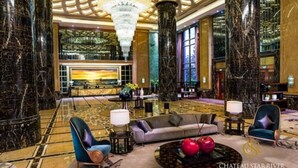 Um lobby luxuoso, com colunas de mármore, um candelabro e uma área de estar com uma mesa redonda e maçãs.