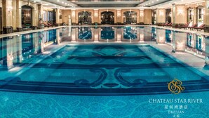 Uma grande piscina interior com design de água azul cristalina, rodeada por espreguiçadeiras e um interior arquitetônico imponente.