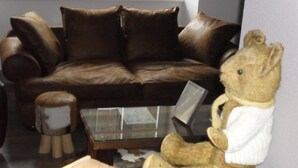 Um sofá de couro marrom com uma mesa de centro de vidro e um banquinho de madeira. Um urso de pelúcia está sentado no chão ao lado de uma caixa com a etiqueta 'Fotos'.