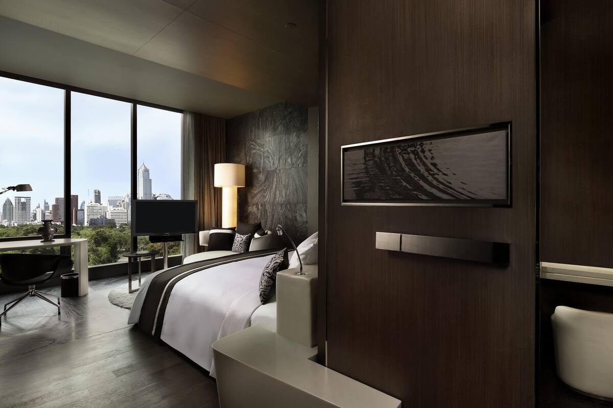 Quarto de hotel moderno com uma janela grande, cama, escrivaninha, cadeira, abajur e vista para a cidade.