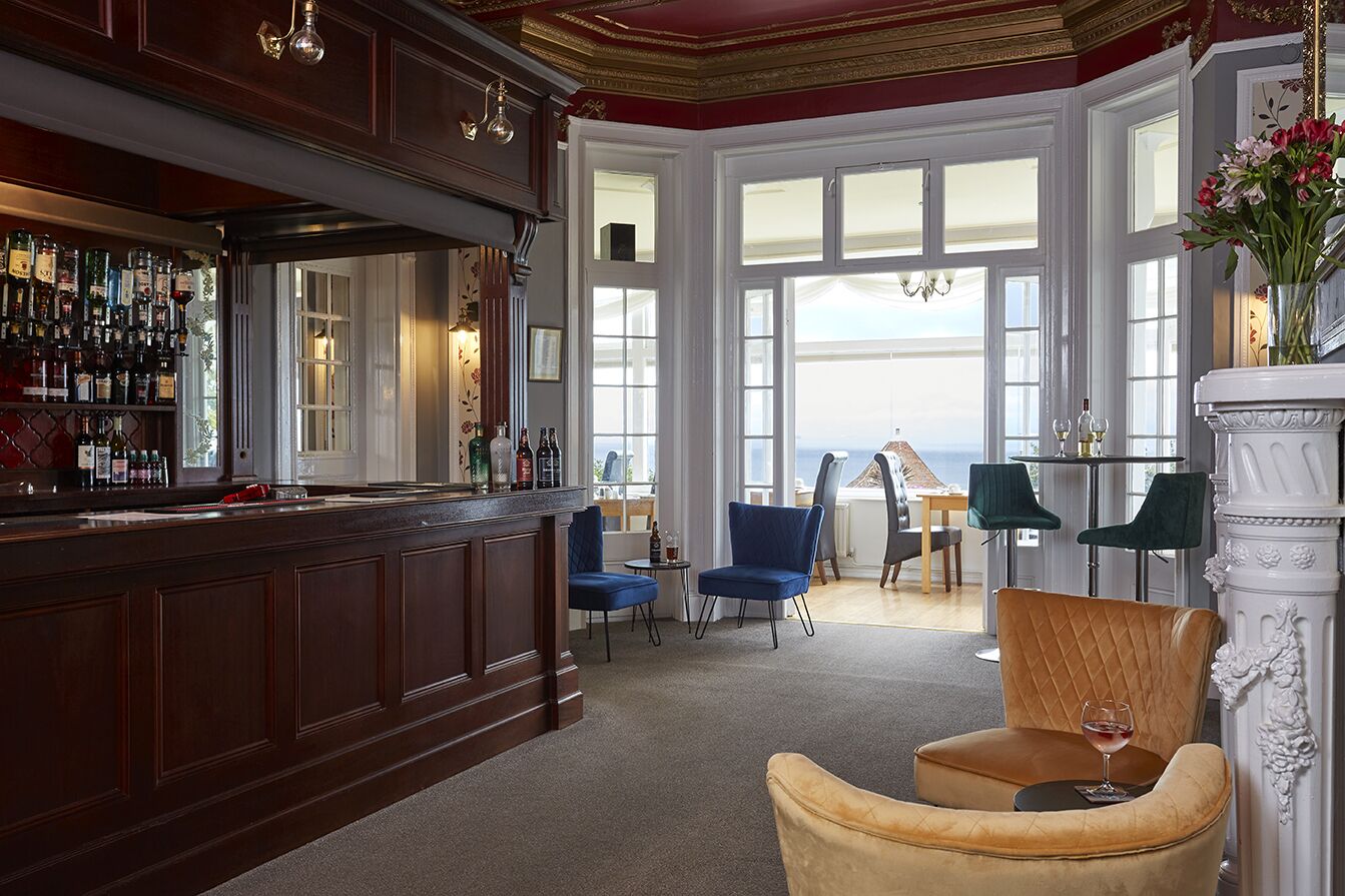 Uma área de bar com uma taça de vinho sobre uma mesa, um balcão com garrafas e vista para o mar através de grandes janelas.