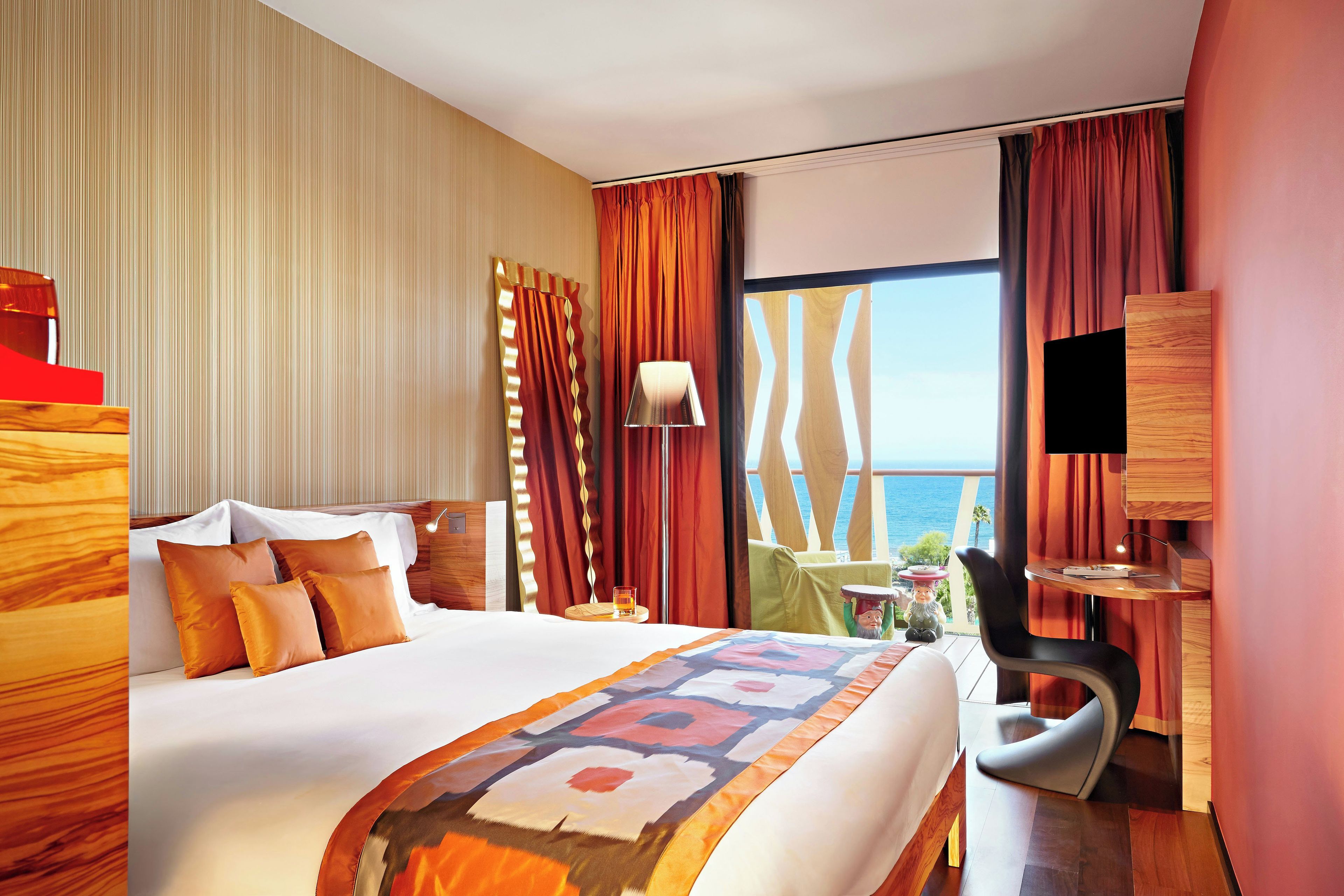 Quarto de hotel com uma cama grande, uma mesa com cadeira, TV na parede e vista para o oceano.