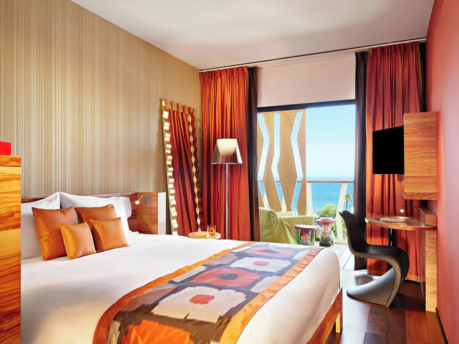 Quarto de hotel com uma cama grande, uma mesa com cadeira, TV na parede e vista para o oceano.