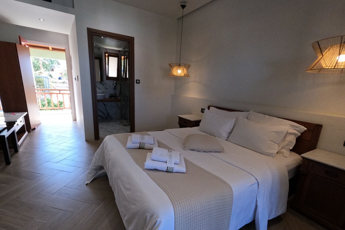 Quarto de hotel com uma cama grande, uma mesa de cabeceira, uma luminária pendente e vista para o banheiro e varanda através de uma porta aberta.