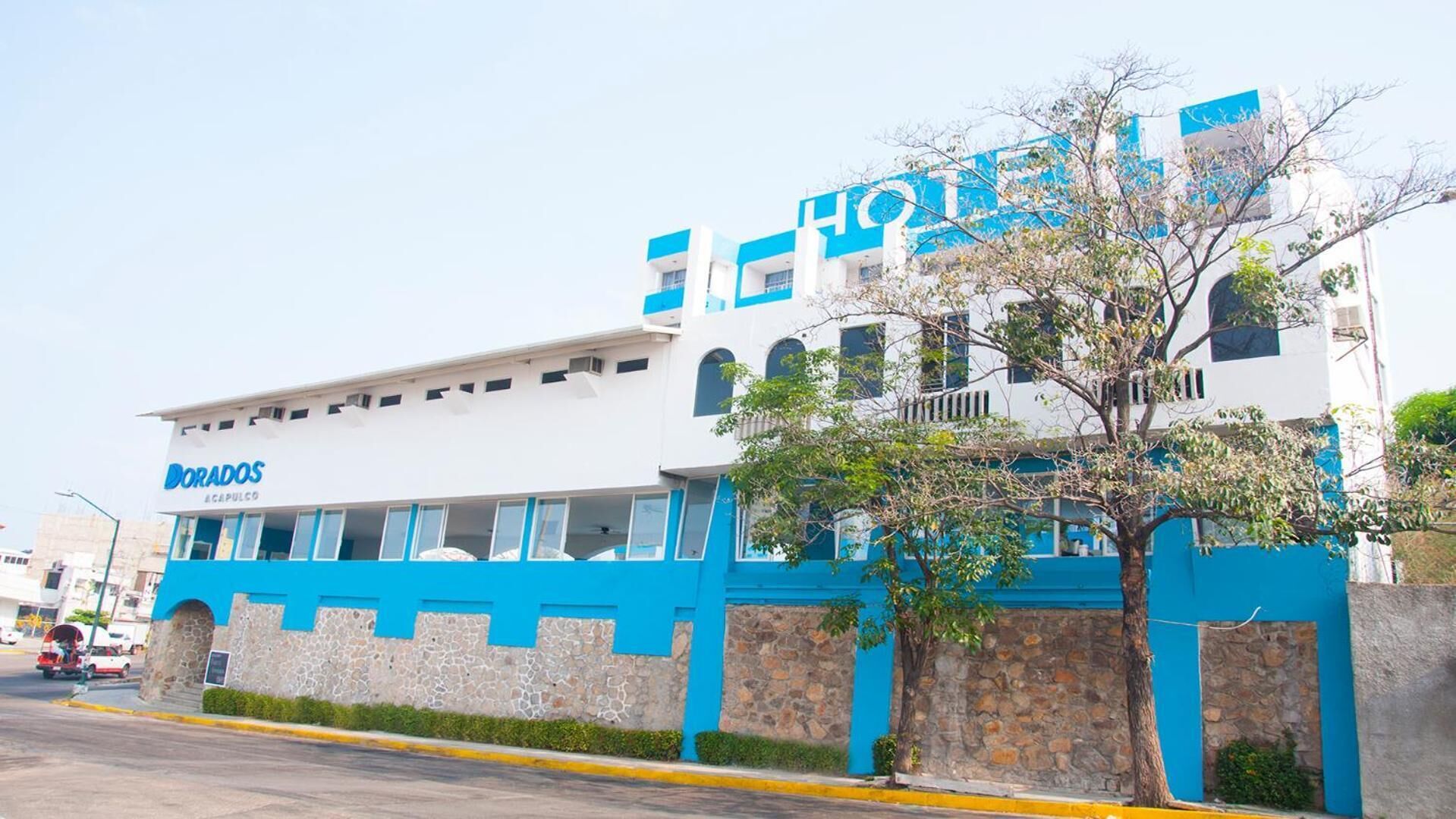 Um hotel com fachada azul e branca, uma placa visível para 'DORADOS' e uma árvore em frente.