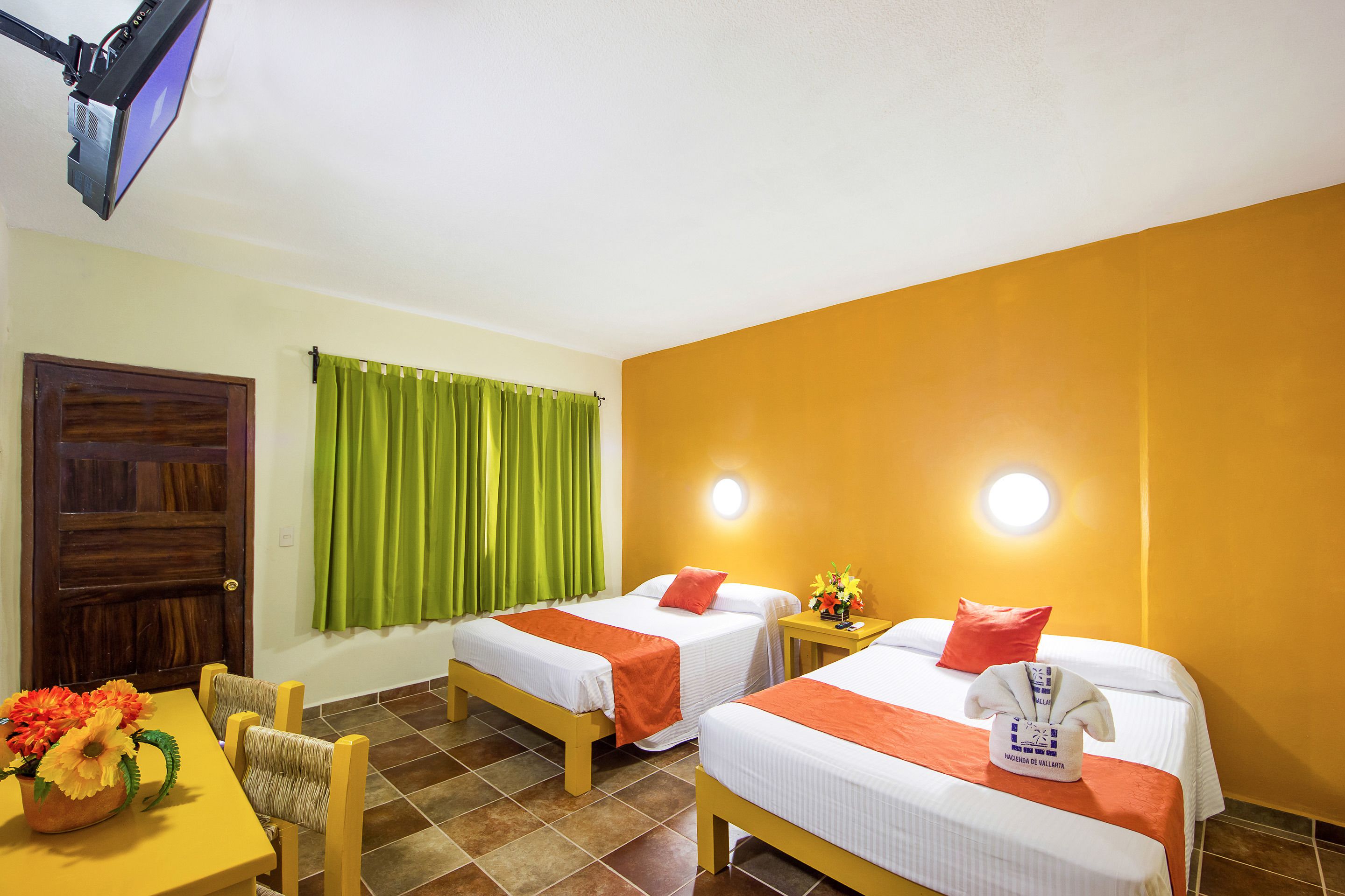 Quarto de hotel com duas camas, uma porta de madeira, cortinas verdes, uma pequena mesa com um vaso de flores e uma luminária na parede.